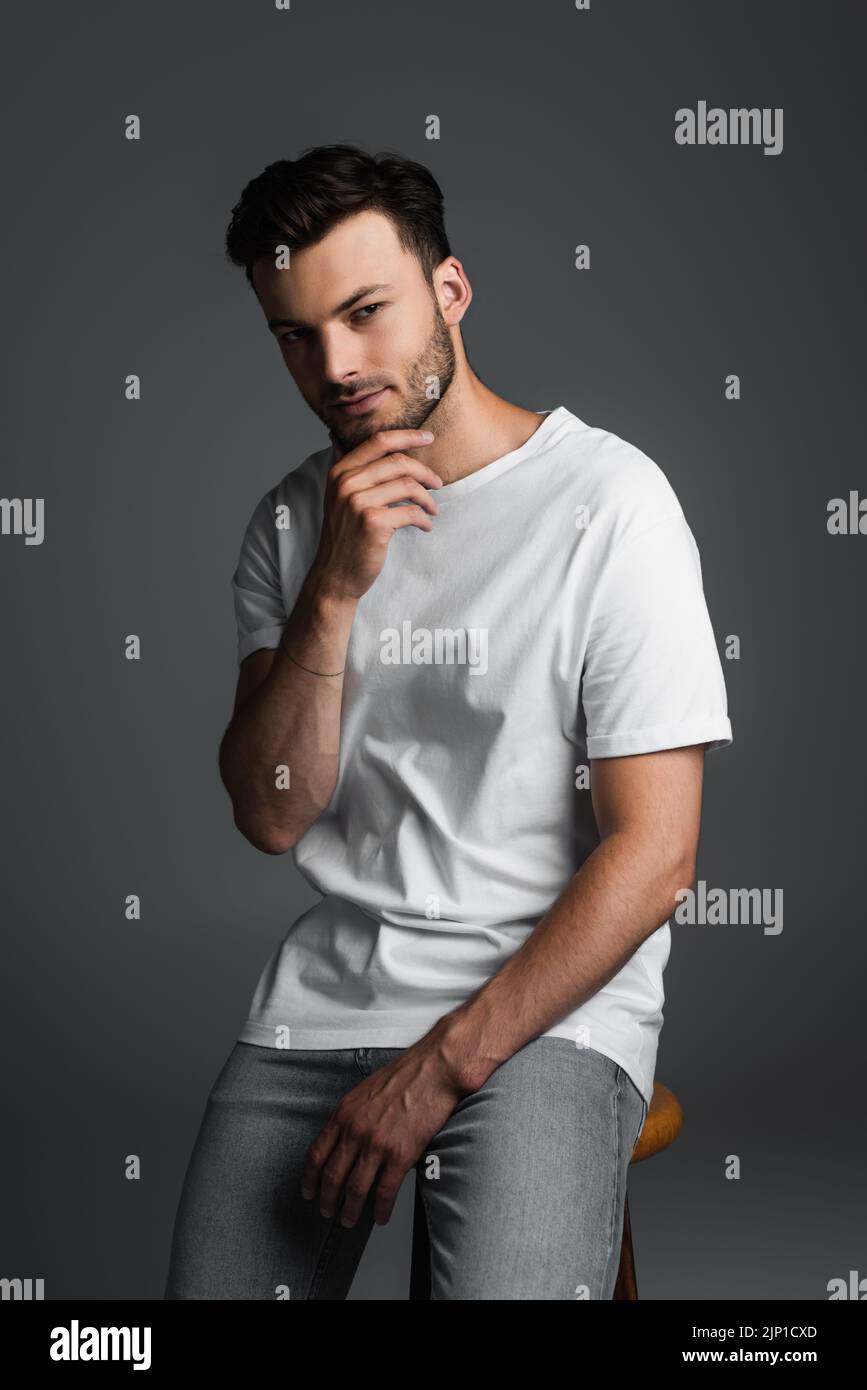 Brunette man in jeans and white t-shirt touching chin while sitting on ...