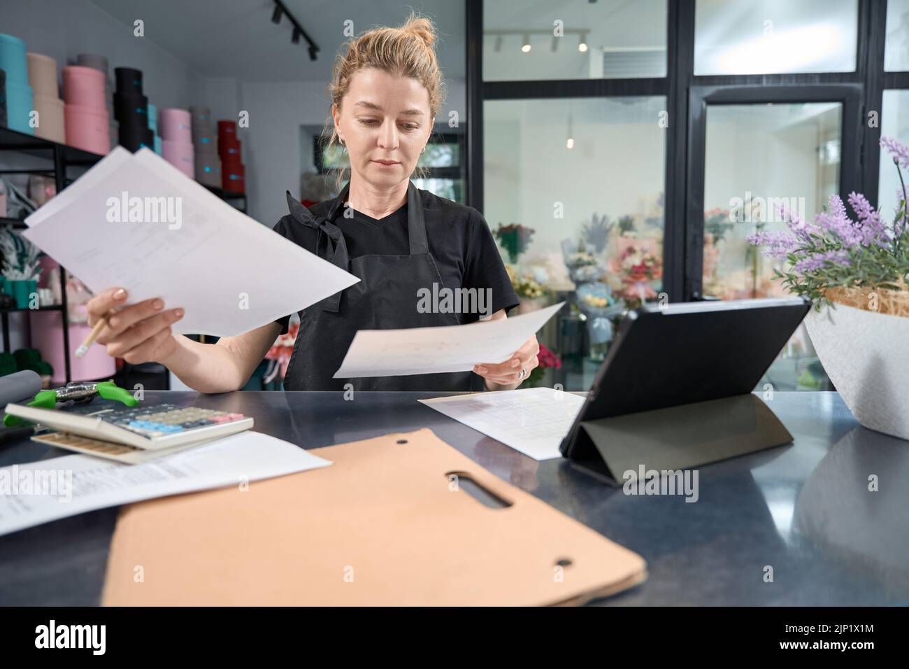 Focused flower shop owner sitting at counter looking through paperwork ...