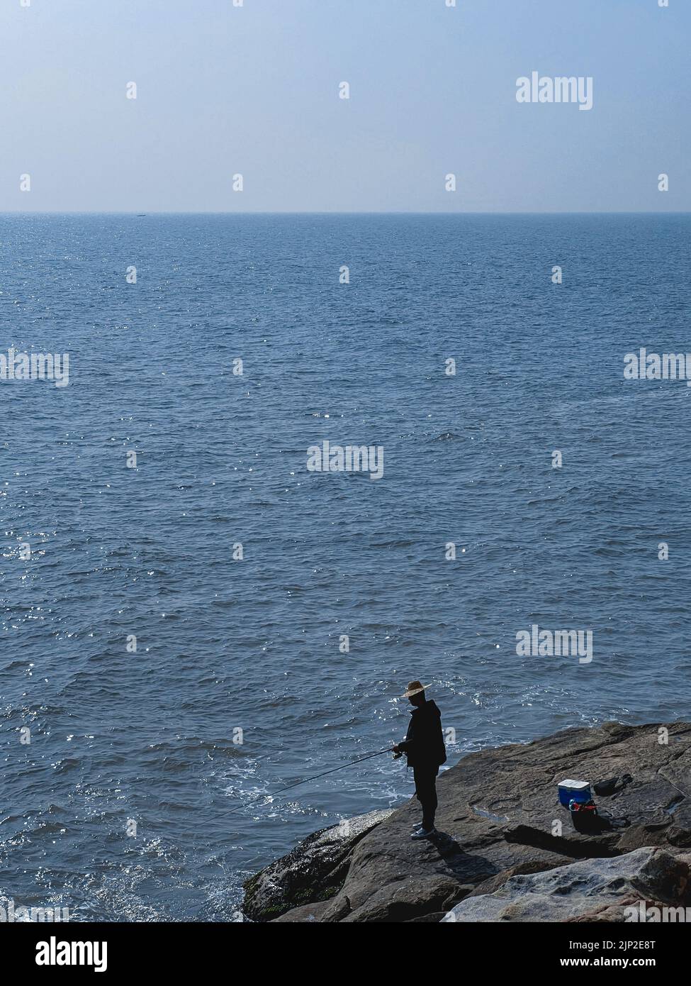 An vertical shot of a person in a hat fishing from a rock near a calm ...
