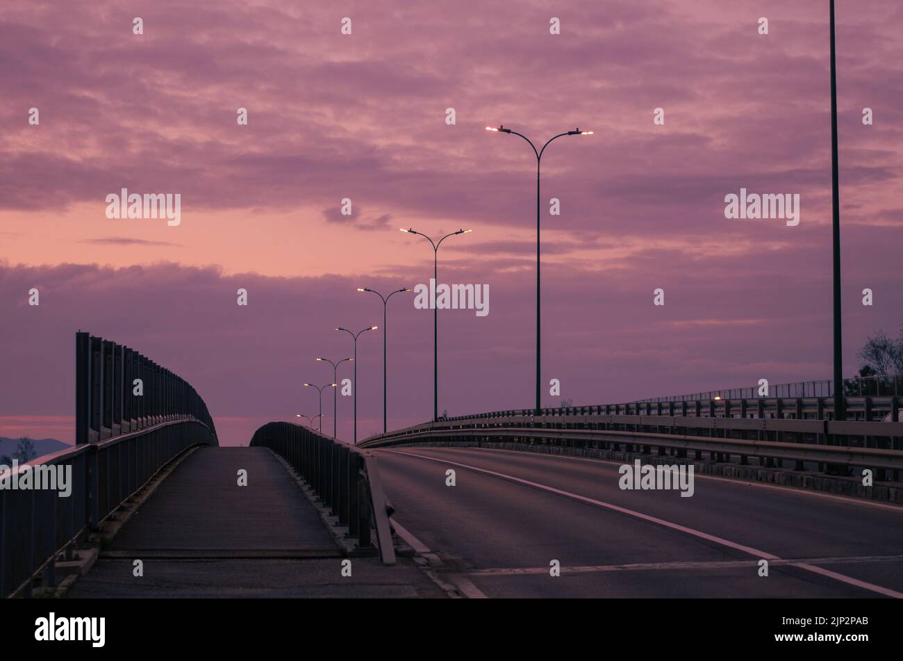 A sunset view of a bridge from the pavement with street lamp posts and ...
