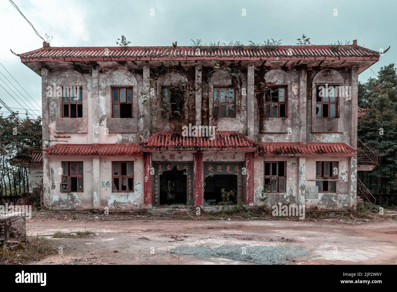 A front view of an old abandoned building with broken windows and plants growing on the walls ...