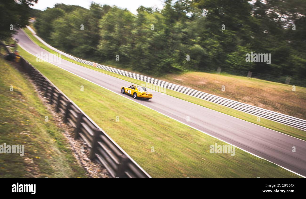 A modern yellow car driving on Brands Hatch racetrack with green trees ...