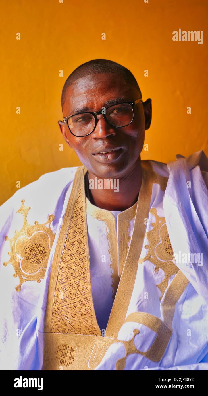 A portrait of an African man against a yellow wall. He wears a typical ...