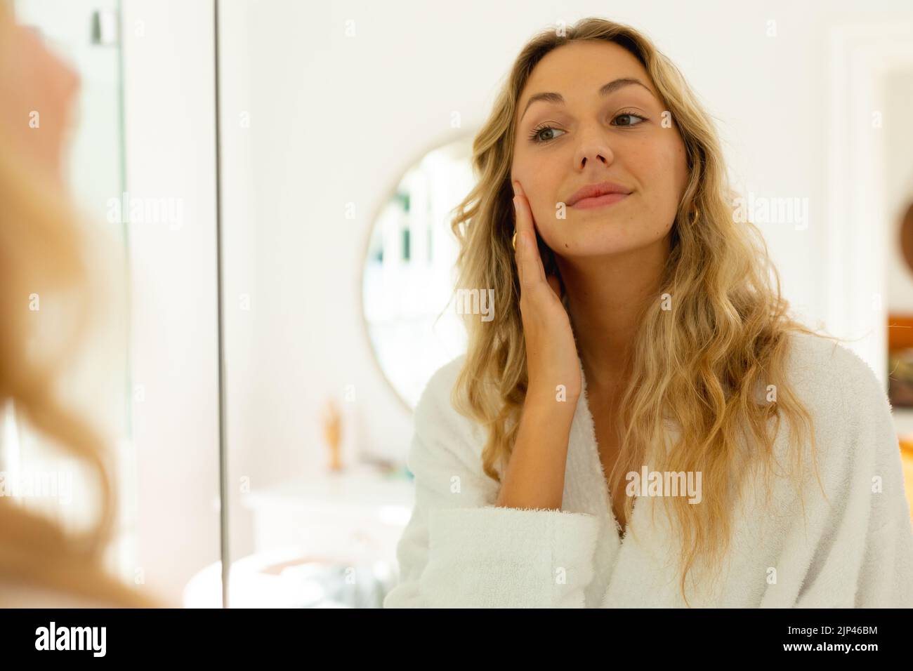 Happy caucasian woman wearing robe, looking at her reflection in mirror ...