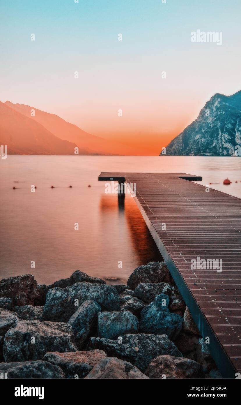 A beautiful view of a calm lake with a wooden dock during orange sunset ...