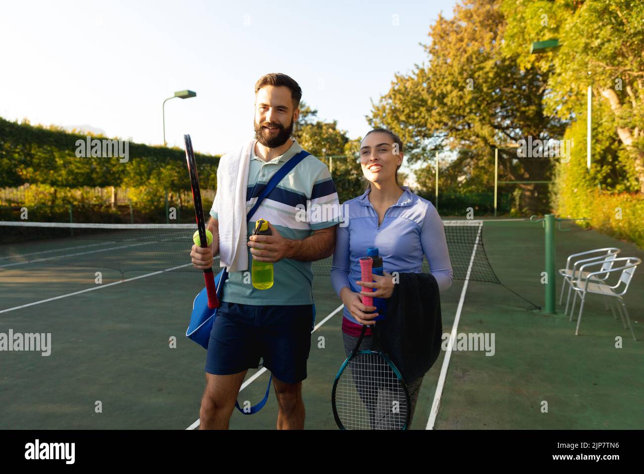 Smiling caucasian couple holding rackets walking on outdoor tennis ...