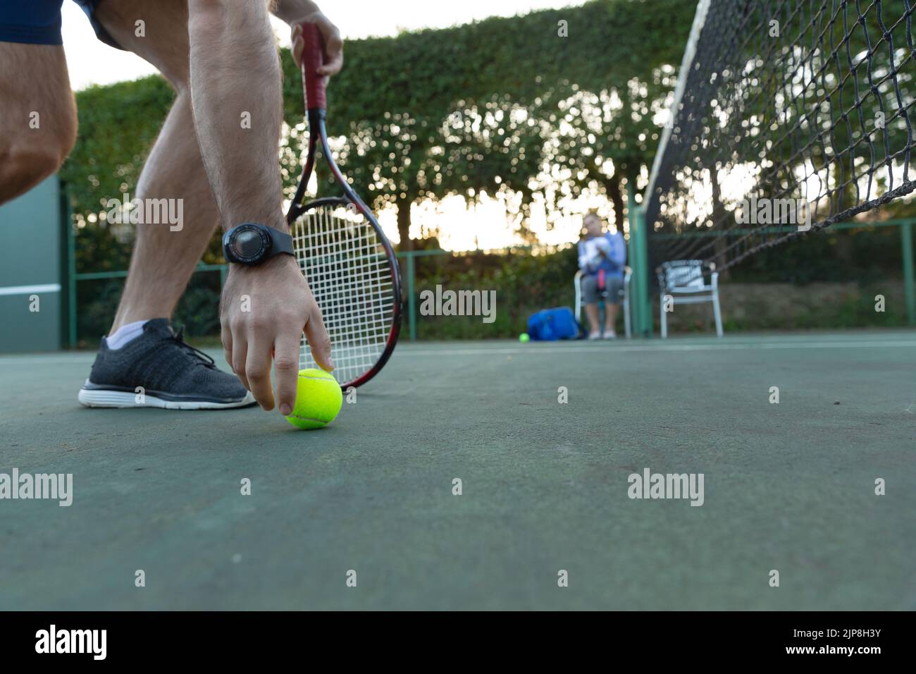 Low section of man holding tennis racket picking up ball on outdoor ...