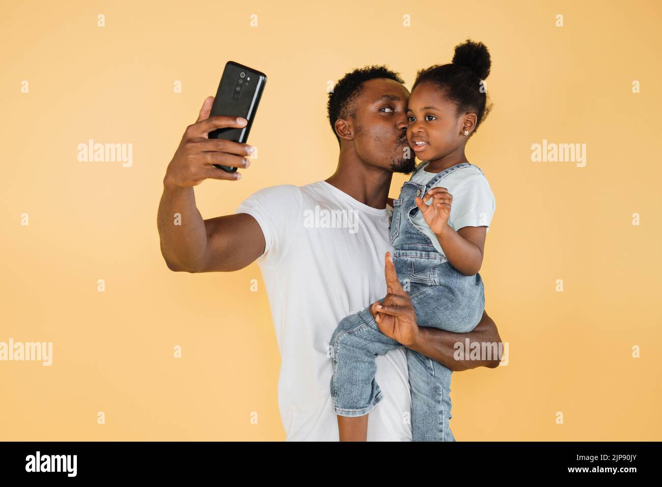 Cheerful african american dad kissing his daughter girl in casual clothes isolated on yellow ...