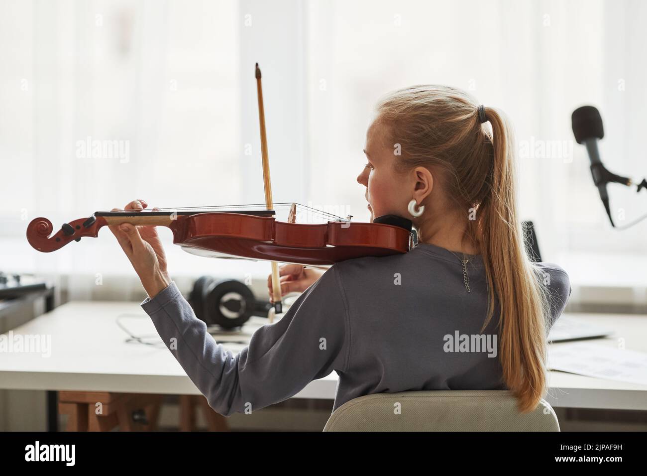 Back view of blonde young woman playing violin in studio against window ...