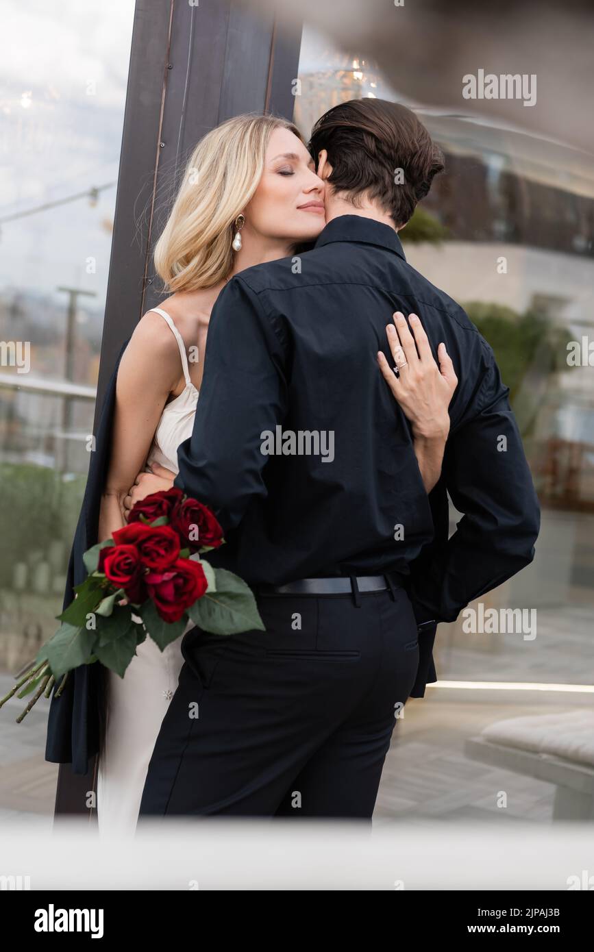 Blonde woman in dress holding red roses and hugging boyfriend outdoors ...