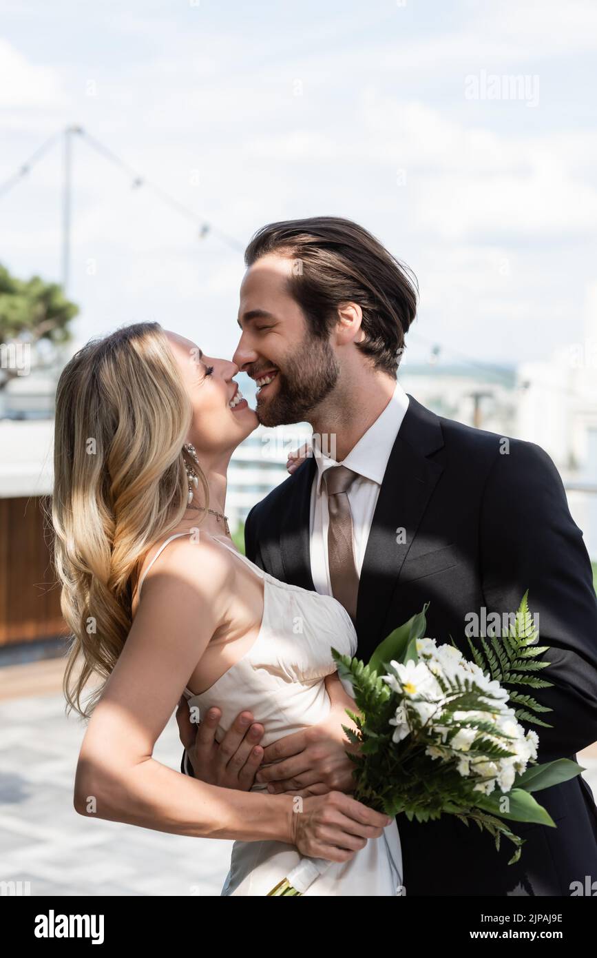 Side view of smiling groom kissing and hugging bride with flowers on ...