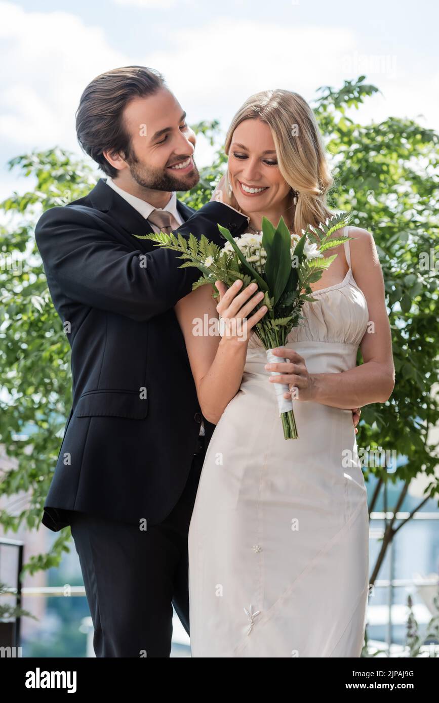 Smiling groom touching bride with bouquet on terrace of restaurant ...
