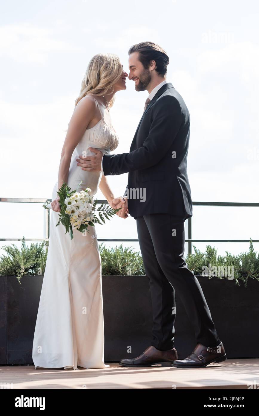Side view of cheerful newlyweds with bouquet kissing and holding hands ...