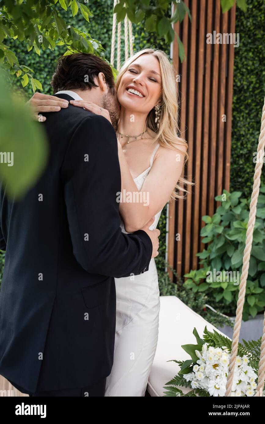 Groom in suit kissing neck of bride near bouquet on terrace of ...
