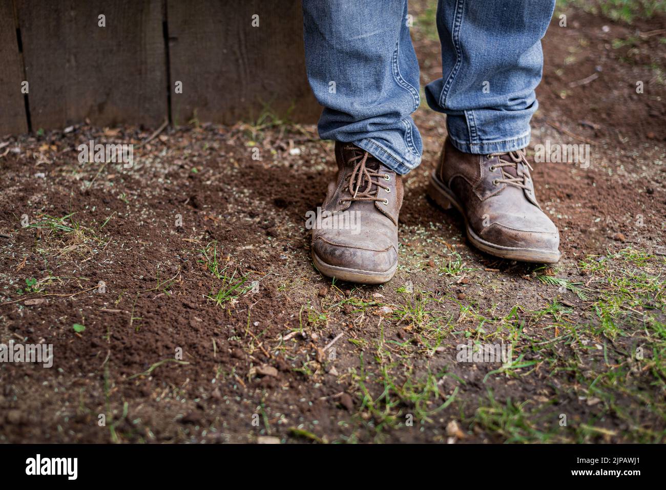 Mans Feet And Lower Legs In Blue Denim Jeans And Brown Work Mans Boots Standing On Patchy