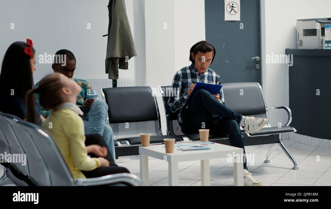 Asian patient filling in medical report at hospital reception lobby, sitting in waiting area to ...