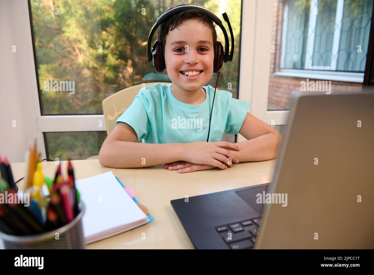 Smart schoolboy in wireless headphones, smiles at camera, sitting at ...