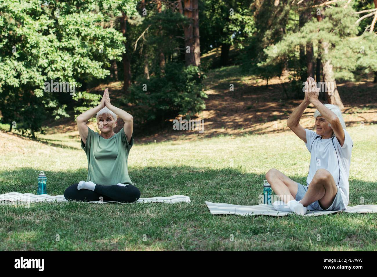 senior couple sitting in yoga pose with praying hands above heads ...