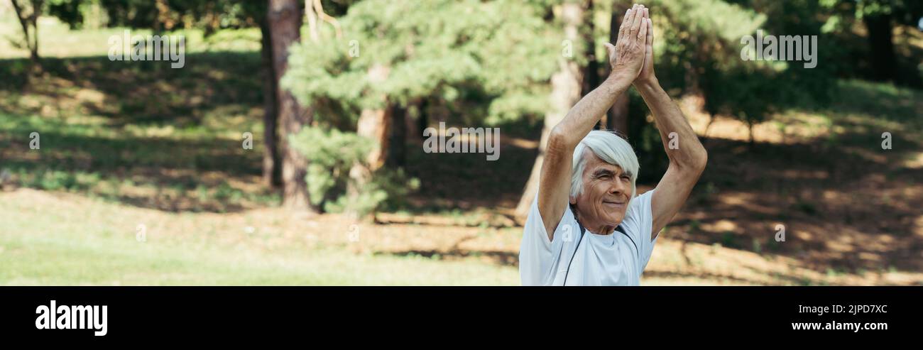 senior man with praying hands above head practicing yoga in green park ...