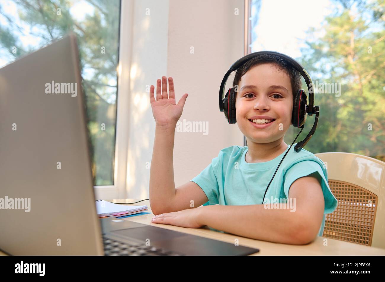 Adorable, smart schoolboy in headphones, waves with hand and smiles at ...