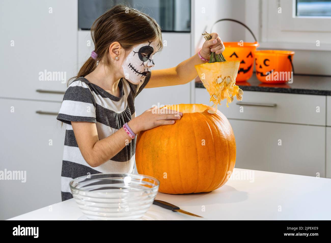 Young girl with skull face paint cutting a pumpkin in their kitchen ...