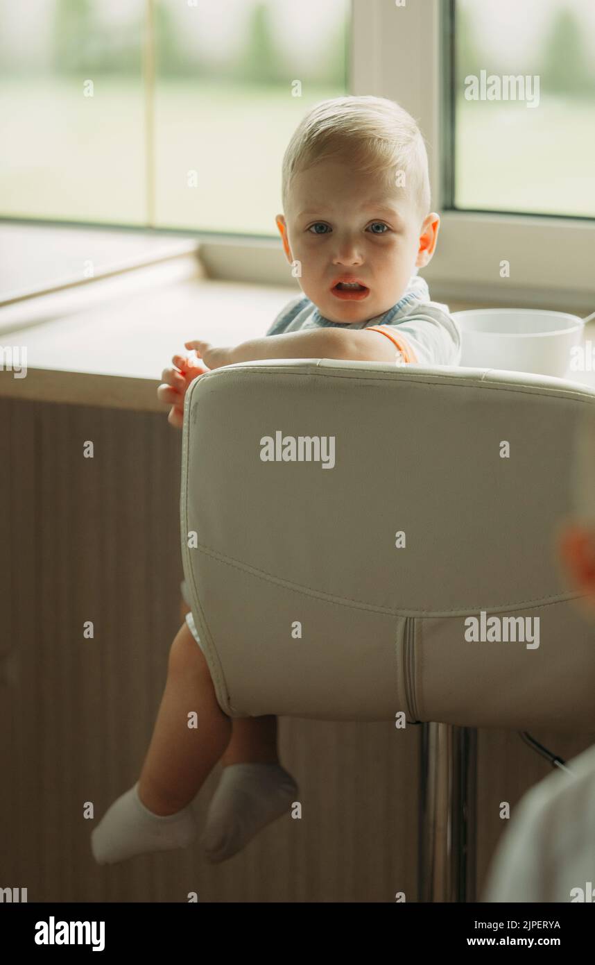 Portrait of a disgruntled little boy sitting on a chair by the window ...