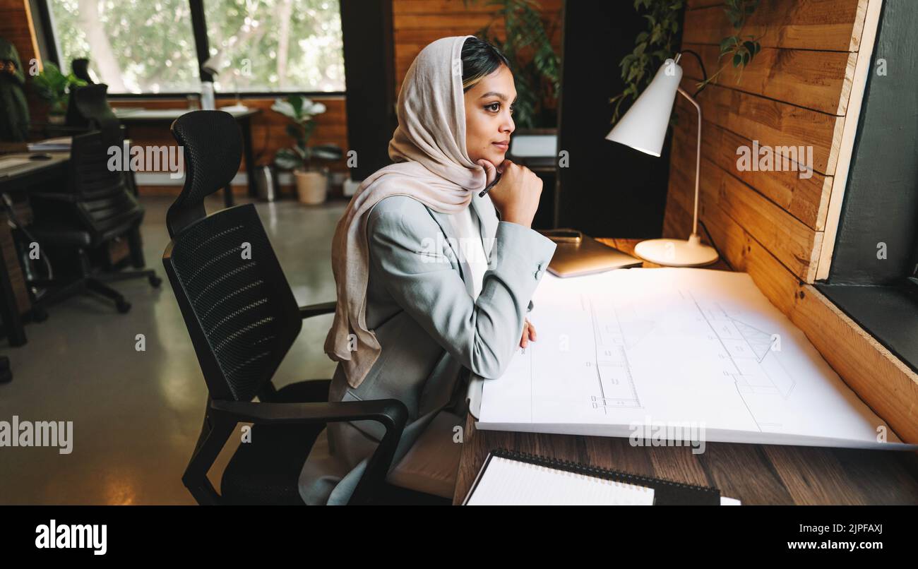 Contemplative Muslim designer working with blueprints in a modern office. Creative young ...