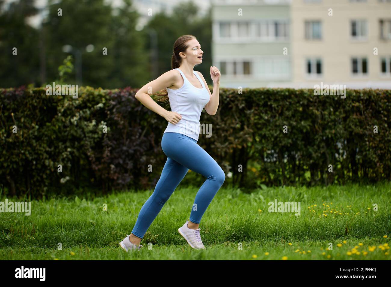 Woman 30s jogging in summer park near residential area with apartment ...