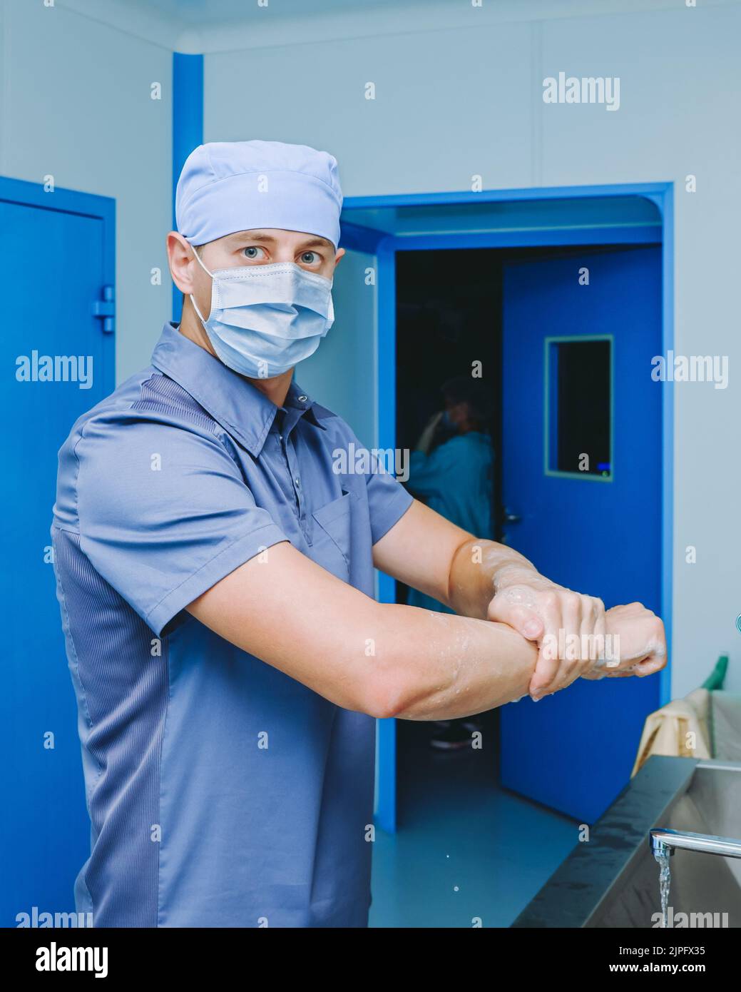 A male doctor in a surgical mask and uniform washes his hands with soap ...