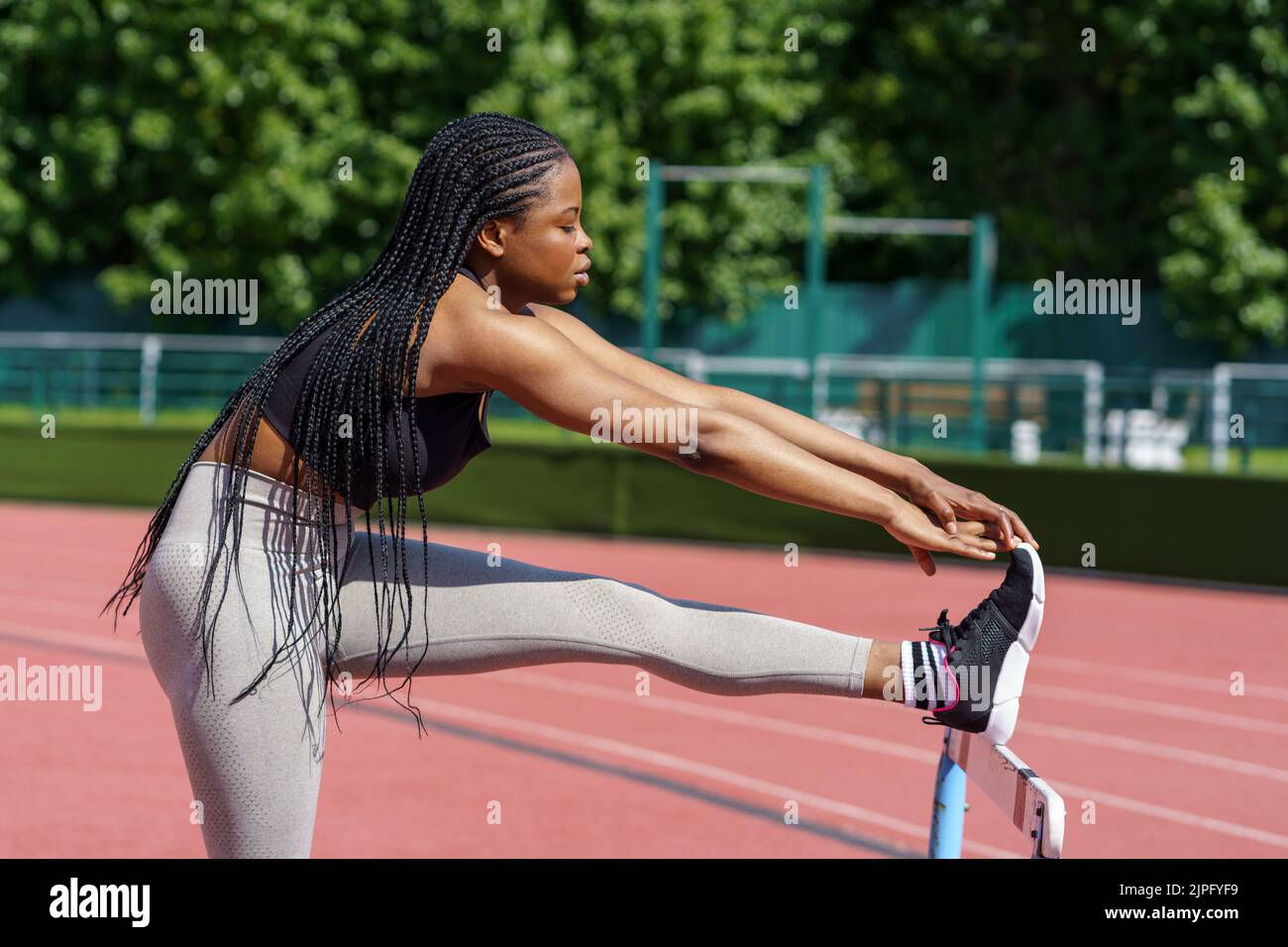 Black young woman with long braids stretches legs and body using hurdles barrier on sports ...