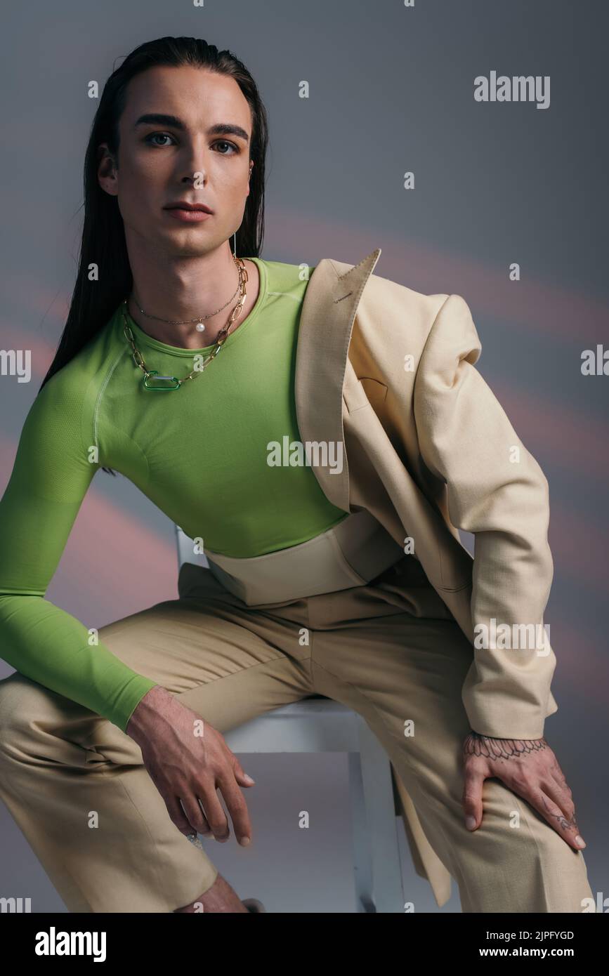Portrait of trendy queer model in beige suit sitting on chair on ...