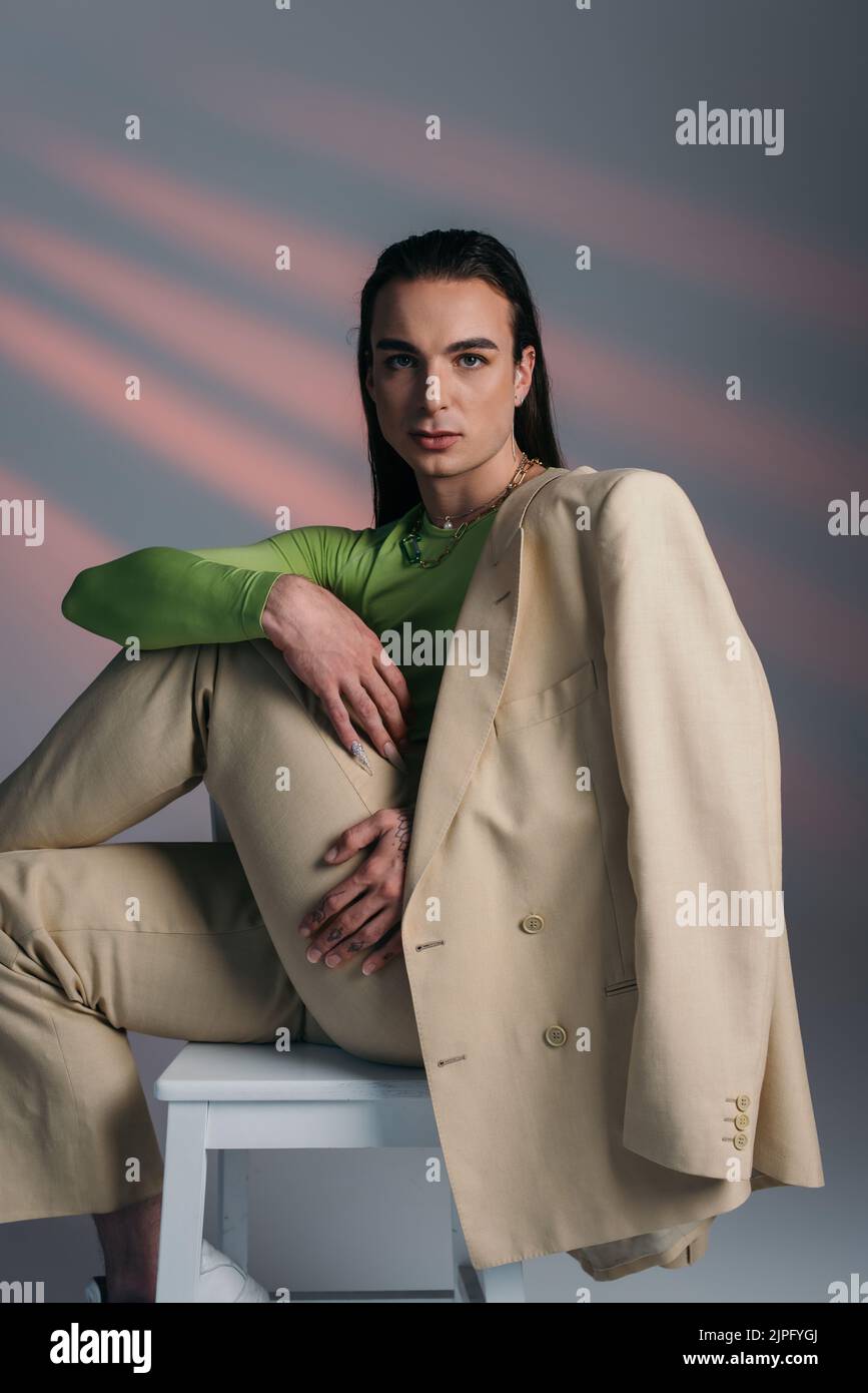 Fashionable queer model in suit posing on chair and looking at camera ...