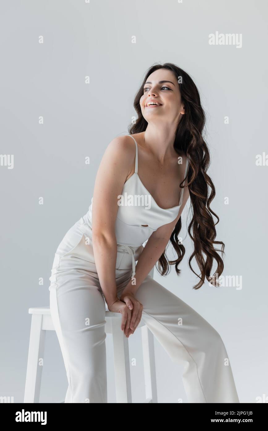 Positive long haired model sitting on chair and looking away isolated ...