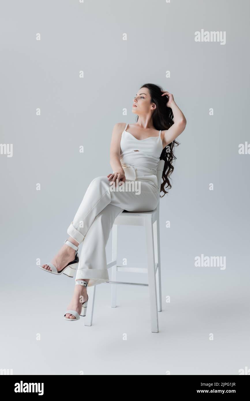 Full length of young model in white clothes and heels sitting on chair ...
