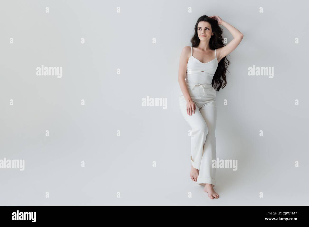 Full length of barefoot woman in white clothes smiling on grey ...