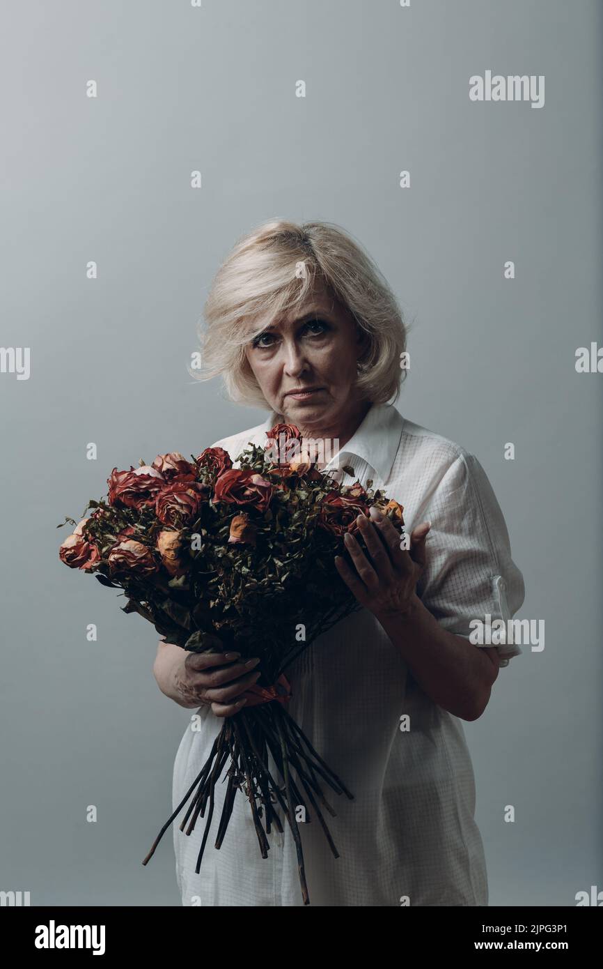 Sad elderly senior woman in white shirt holds withered one dry old dead ...