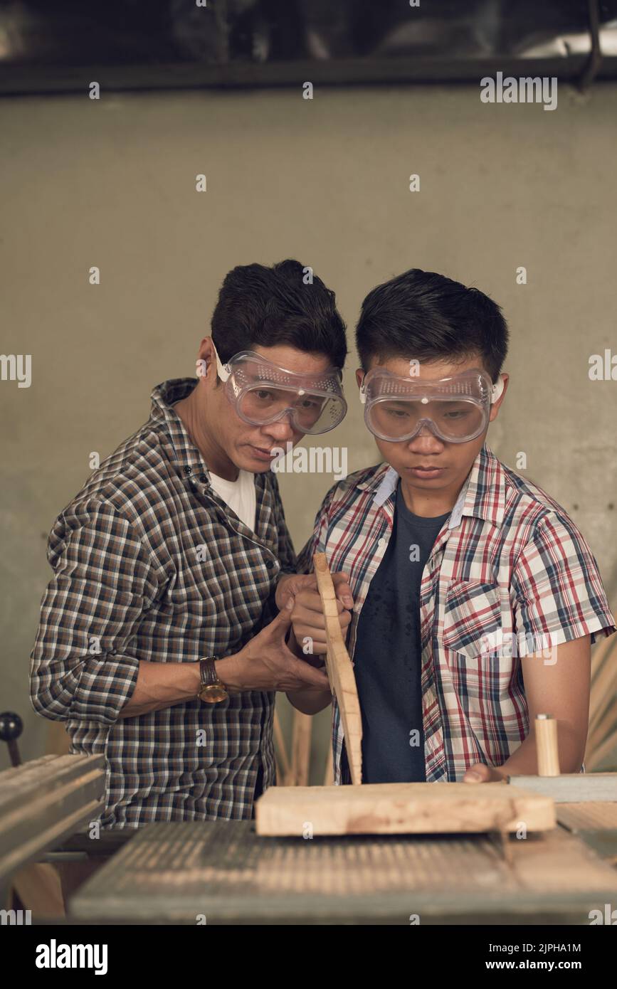 Middle-aged Vietnamese man explaining his teenage son how to saw wood ...
