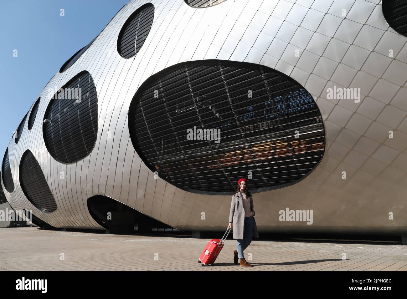 young woman tourist with a red travel suitcase by the futuristic facade ...