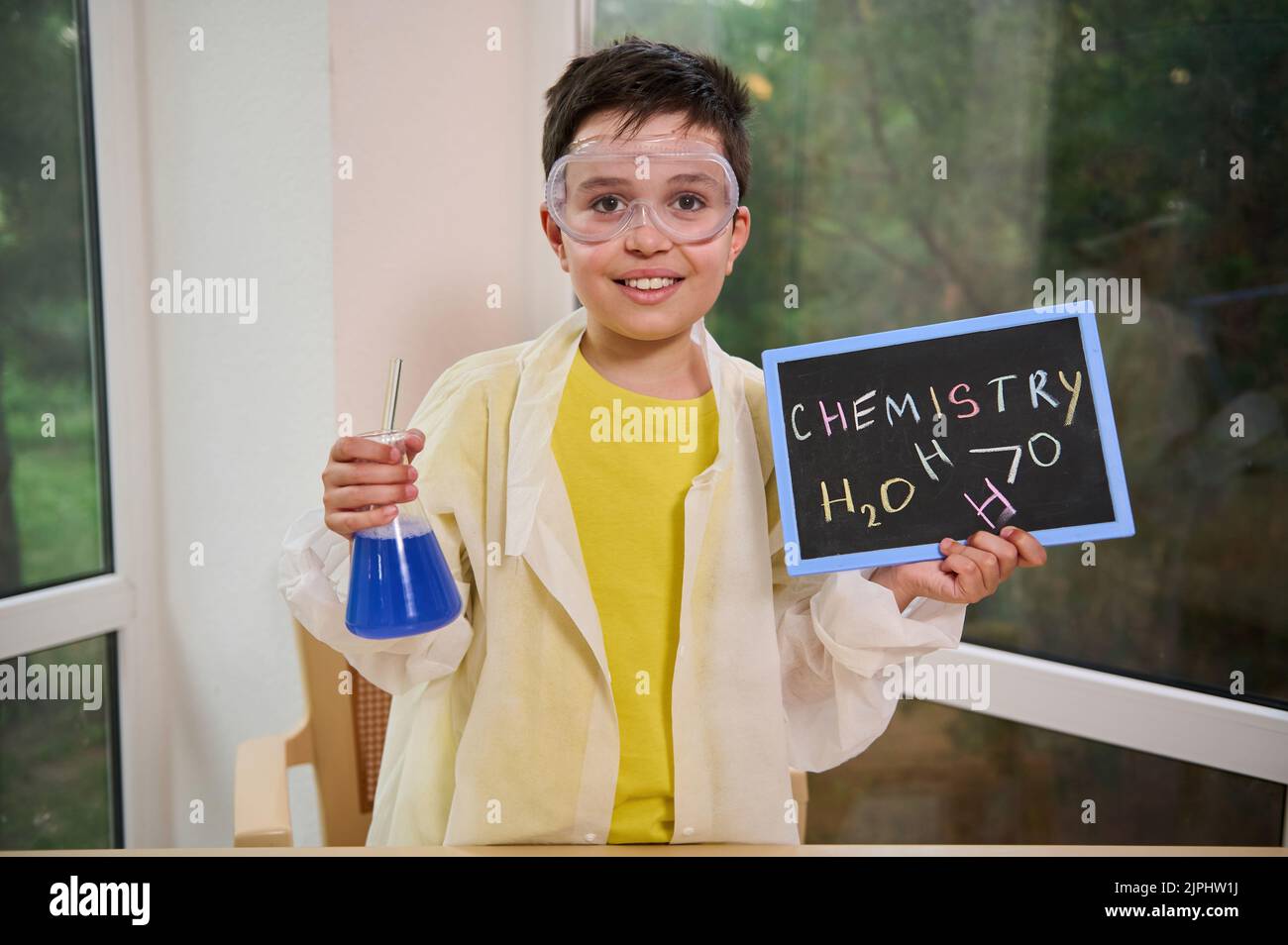Little chemist in safety lab wear, holding flask with blue chemical ...