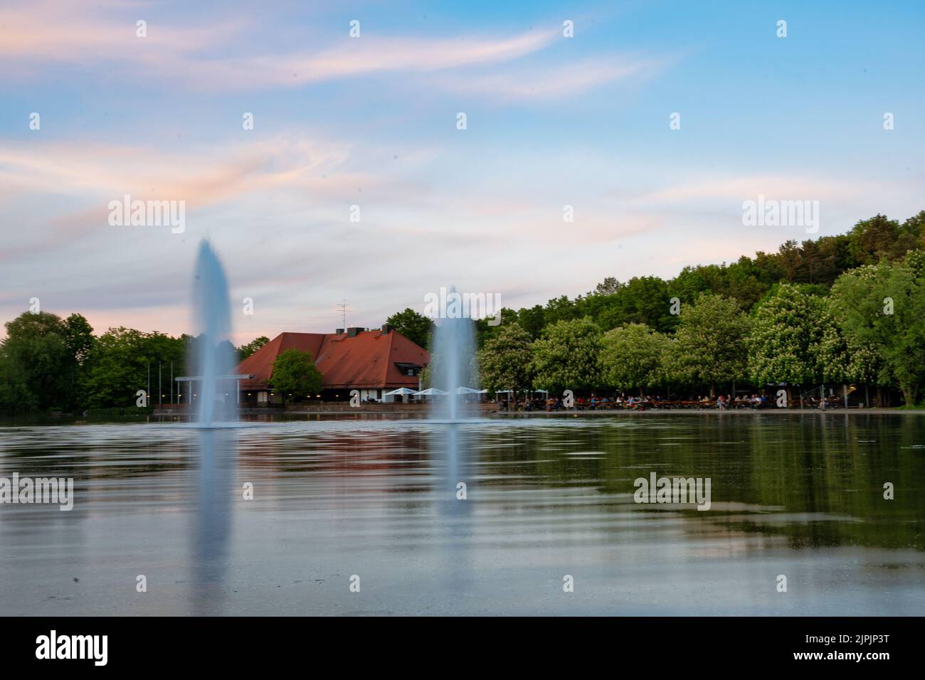 A lake with 2 fountains in front of a restaurant with many trees Stock ...
