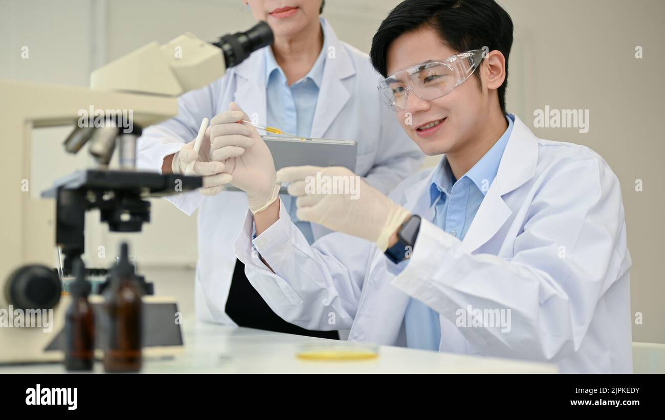 Handsome and talented young Asian male scientist in goggles and gown working in the laboratory ...