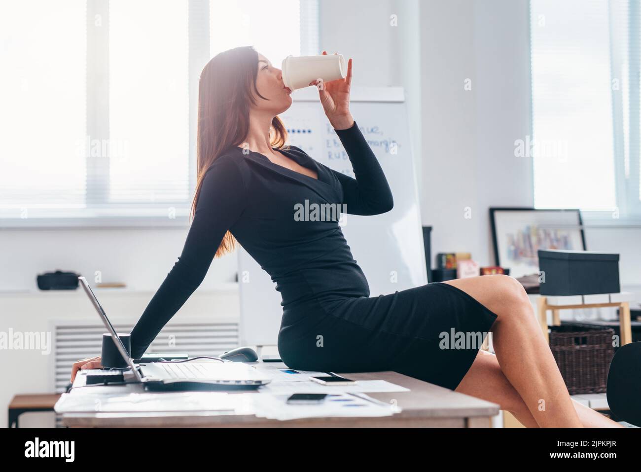 Woman in an office sitting on her desk drinking coffee Stock Photo - Alamy