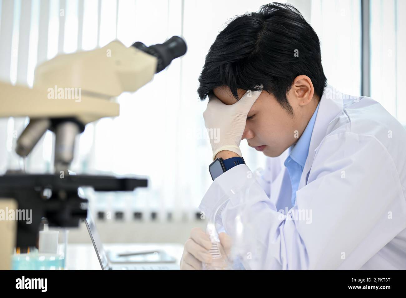 Stressed young Asian male scientist or medical technician in white gown ...