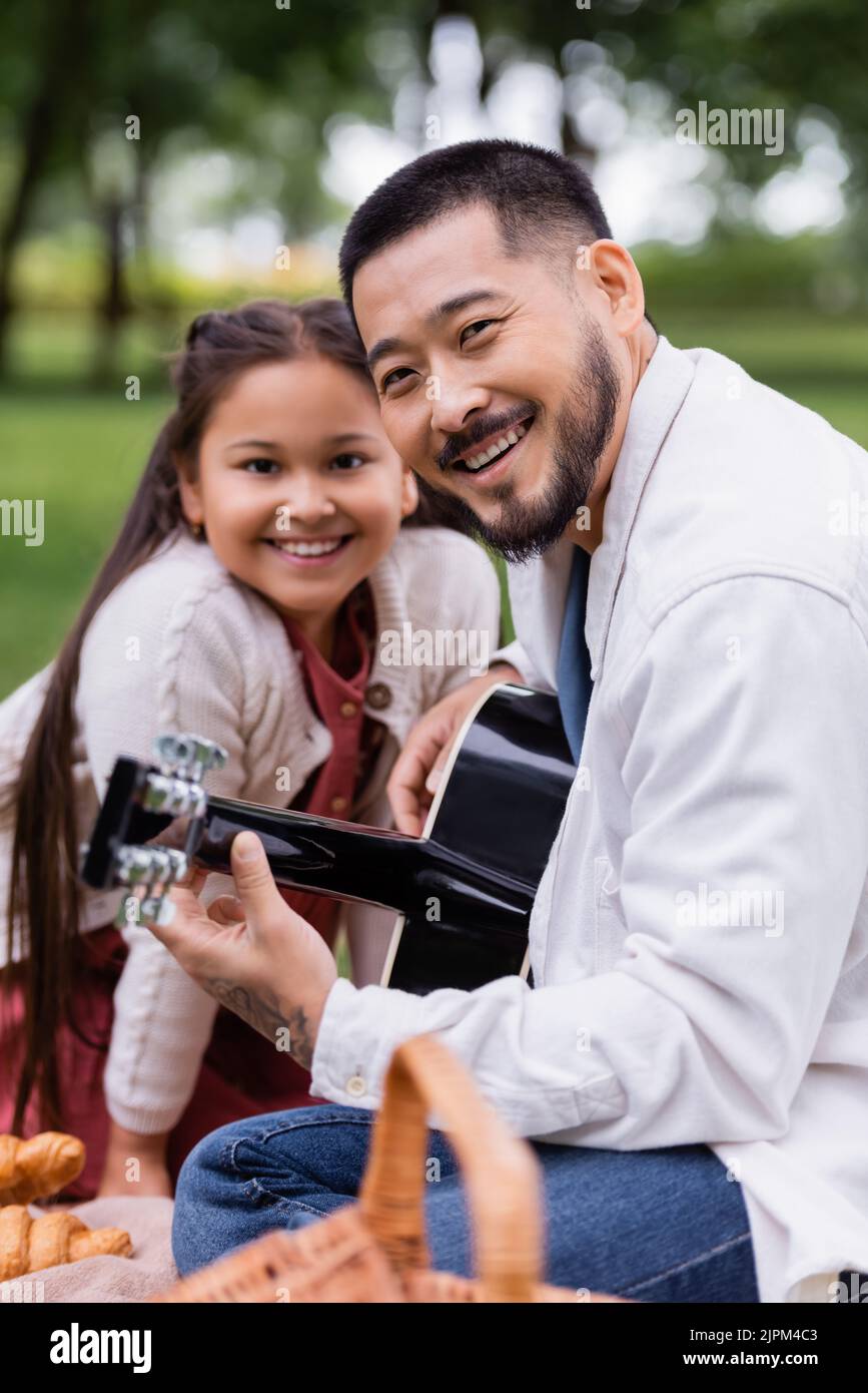 Positive asian man playing acoustic guitar and looking at camera near ...