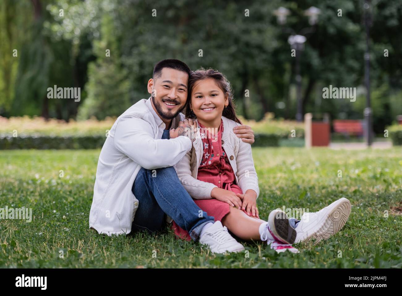 Positive asian man hugging daughter while sitting on grass in summer ...