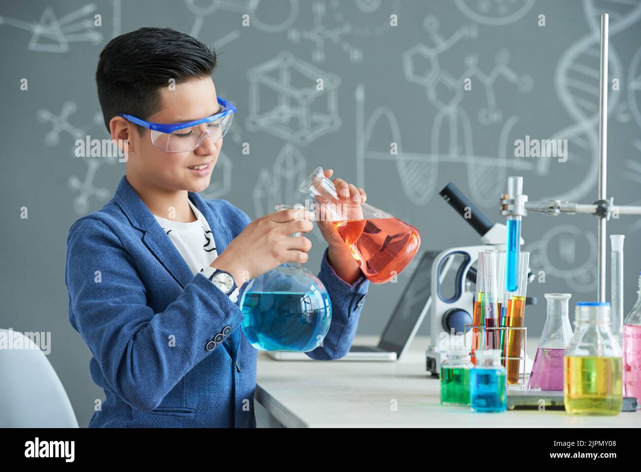 Gifted young researcher wearing safety goggles pouring red liquid into ...