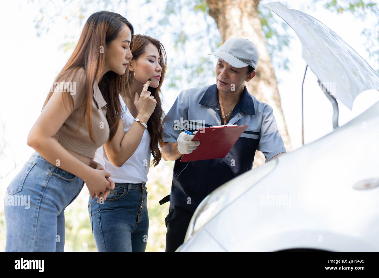 Asian male auto mechanic holds a clipboard and examines car engine ...