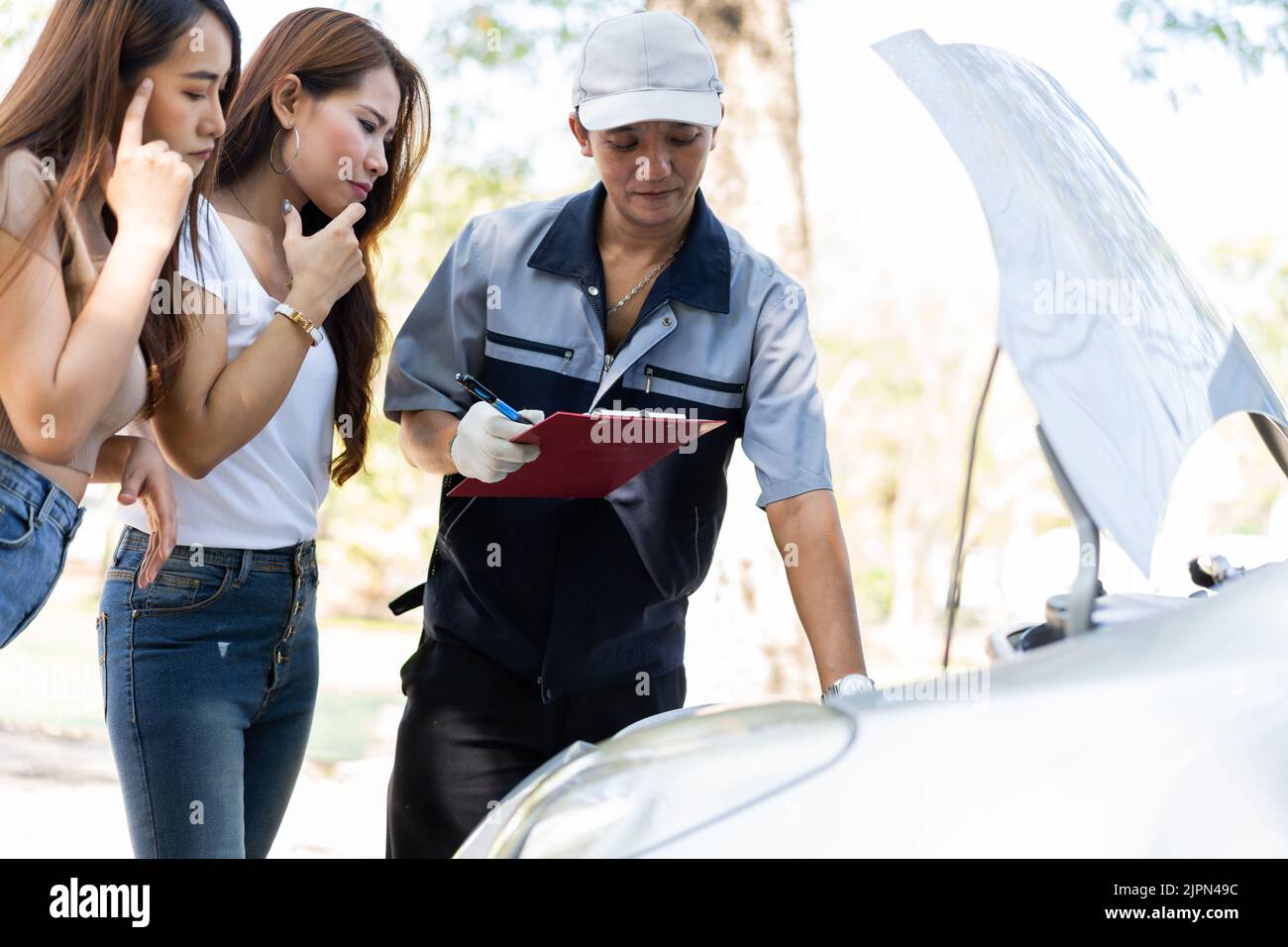 Asian male auto mechanic holds a clipboard and examines car engine ...