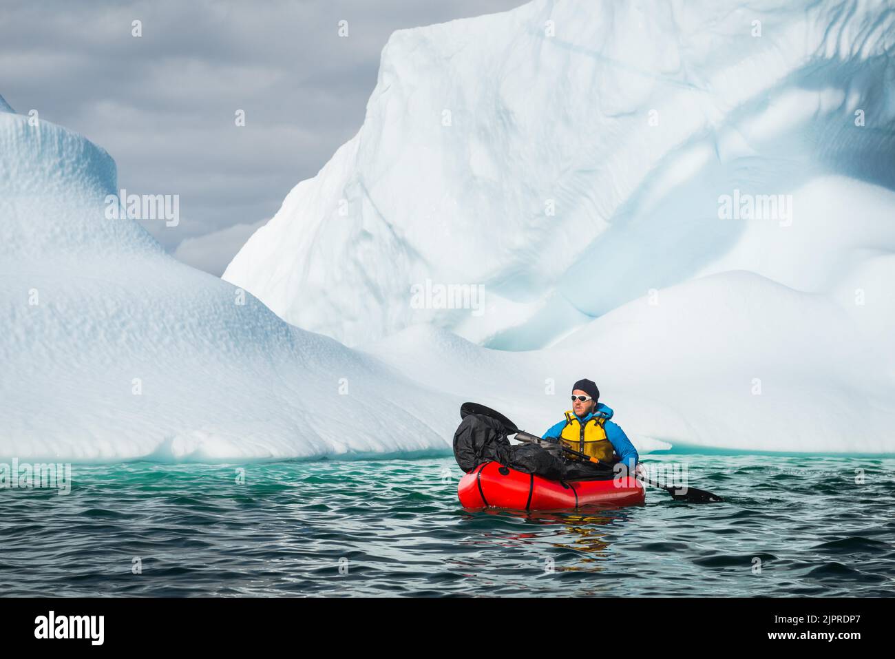 Man with packraft on fjord, icebergs and mountains behind, cloudy ...