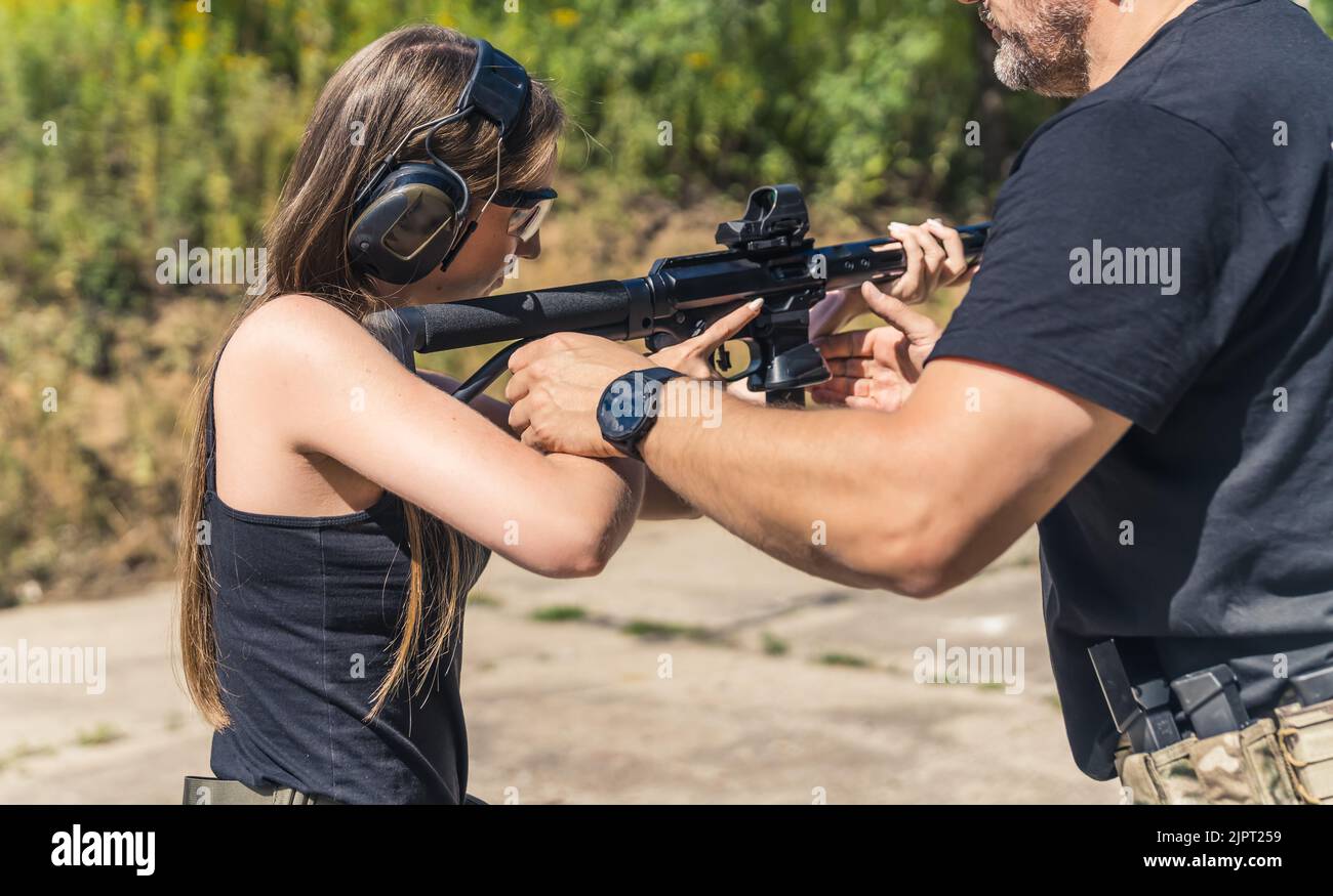 Male instructor helpin female client wearing safety goggles and headphones use submachine gun ...